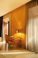 Quarto de hotel com secretária de madeira, uma cadeira, um candeeiro e uma janela grande com cortinas.