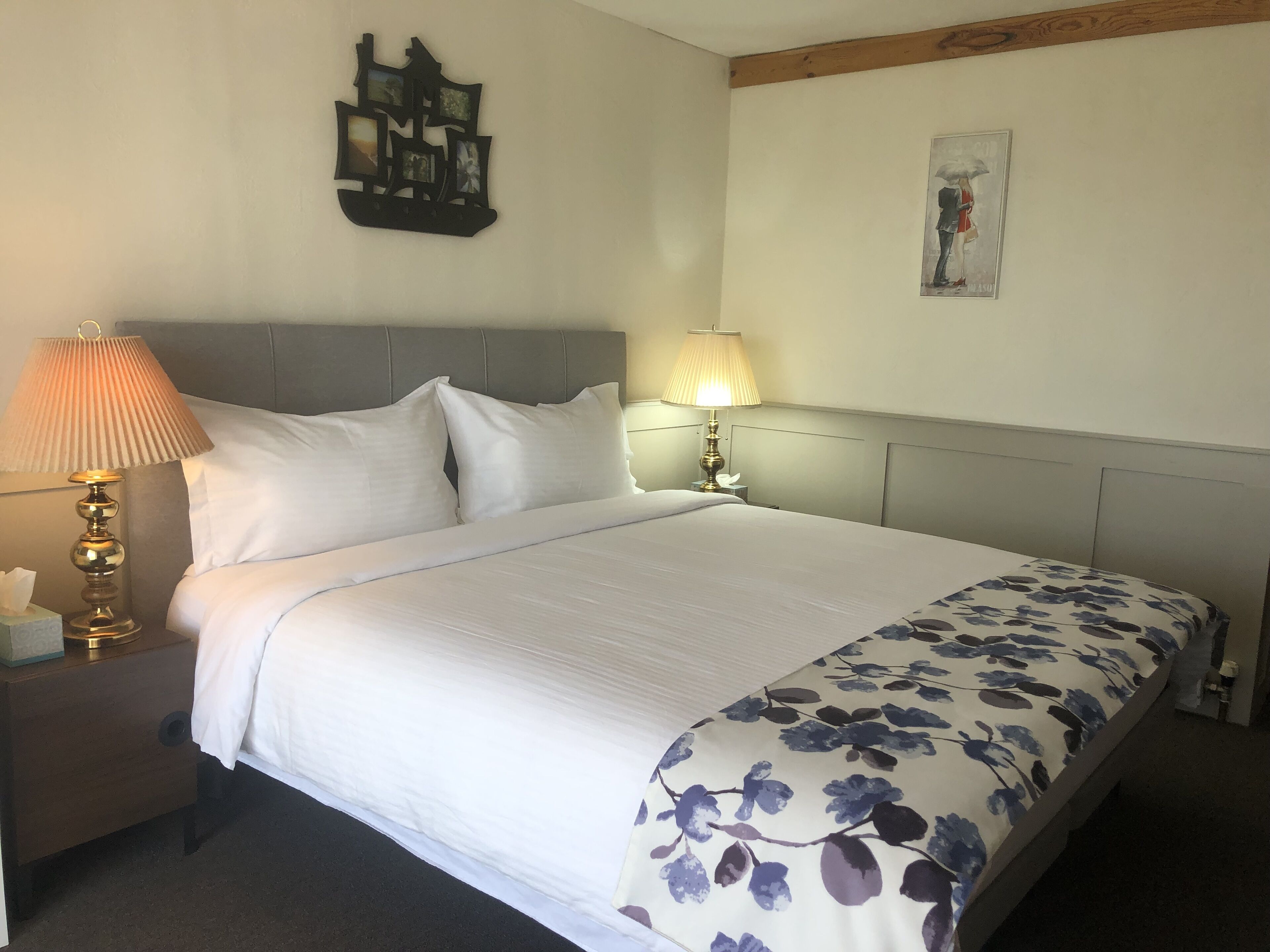 Cama bem arrumada com lençóis brancos e manta com padrão floral. Há duas mesas de cabeceira, cada uma com uma luminária e uma caixa de lenços. Um enfeite de parede em formato de navio e um quadro estão visíveis na parede.