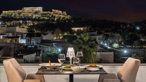 Mesa de jantar com pratos, copos e um abajur, com vista para uma paisagem urbana histórica com ruínas antigas.