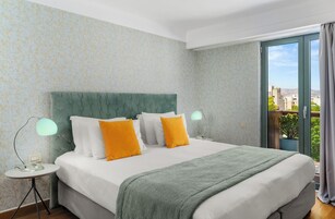 Quarto de hotel com cama, mesa de cabeceira, luminária e vista da cidade por uma janela ampla.