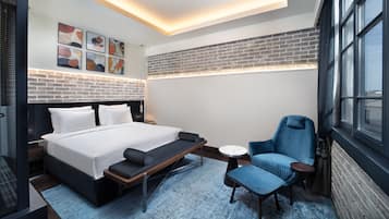Quarto de hotel moderno com uma cama grande, uma poltrona azul, uma mesa redonda pequena e uma parede de tijolos decorativa.