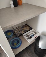 Uma bancada de cozinha com cafeteira, lixeira e área de armazenamento com pratos e copos.