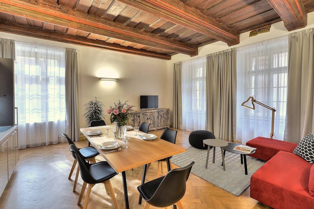 Uma sala de jantar com uma mesa de madeira posta para quatro pessoas, cadeiras pretas, um sofá vermelho e uma televisão sobre um móvel de madeira.