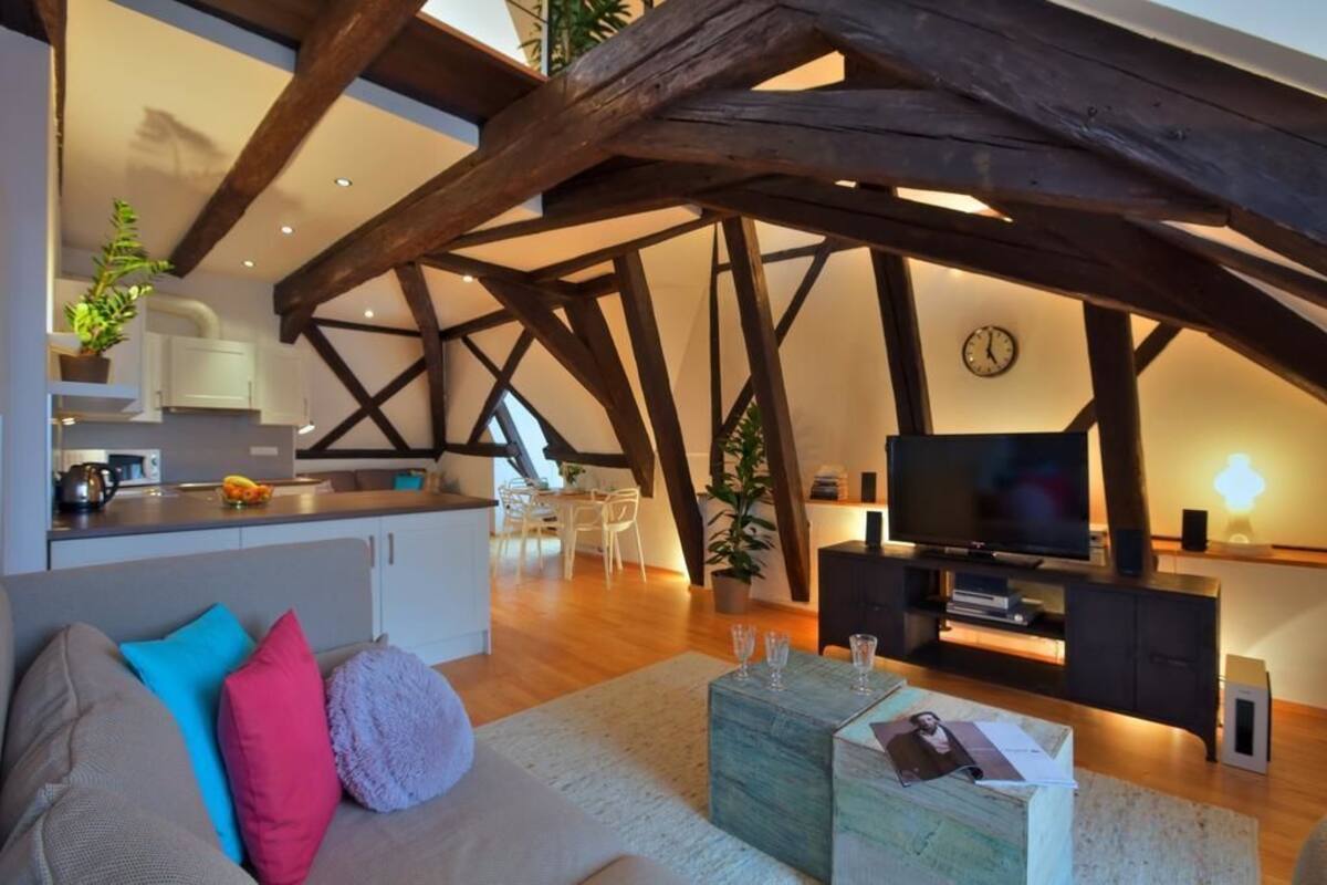 Um loft moderno com vigas de madeira aparentes, área de cozinha, sala de estar com sofá e mesa de centro, e uma televisão.