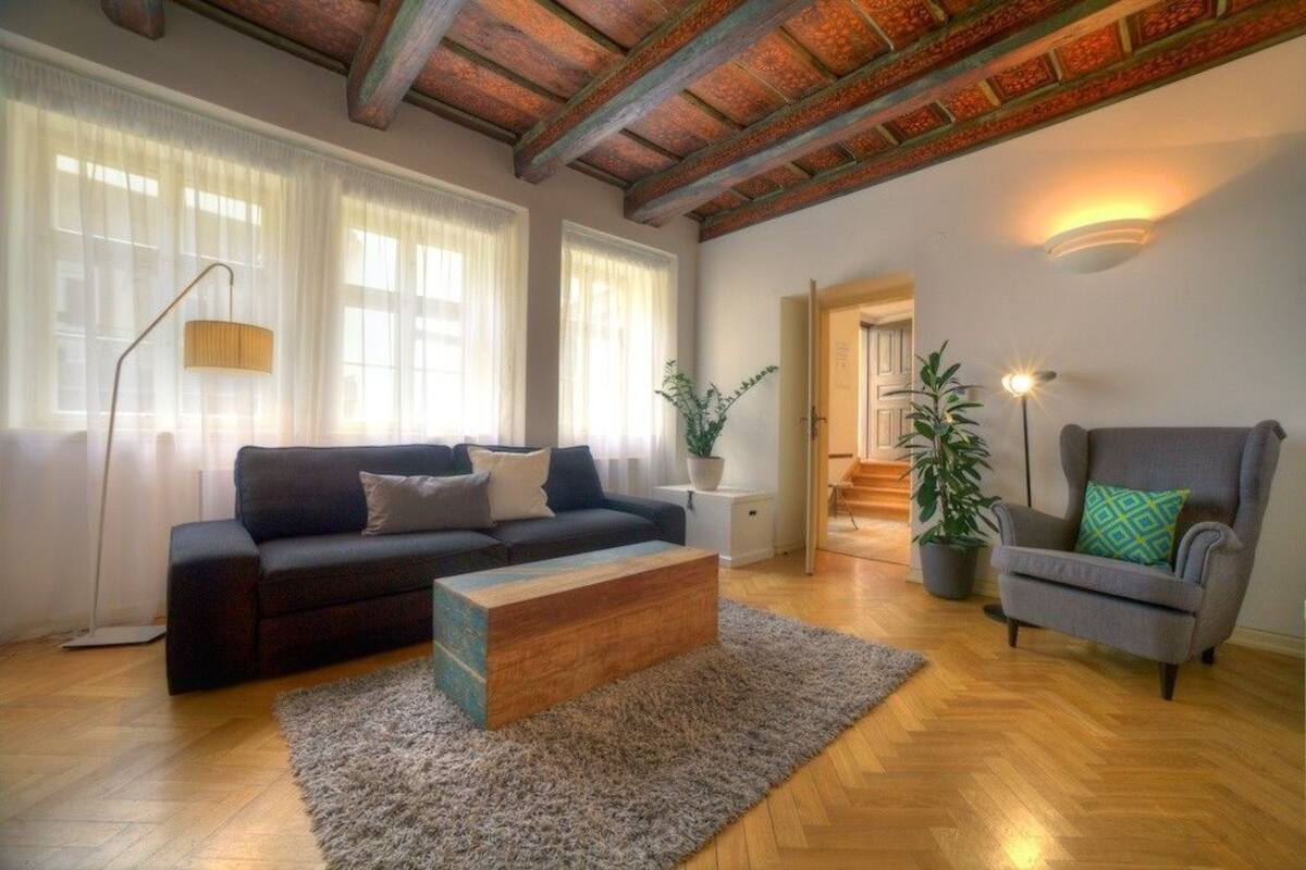 Uma sala de estar com um sofá escuro, uma mesa de centro de madeira, um tapete cinzento, um abajur e uma planta em vaso.