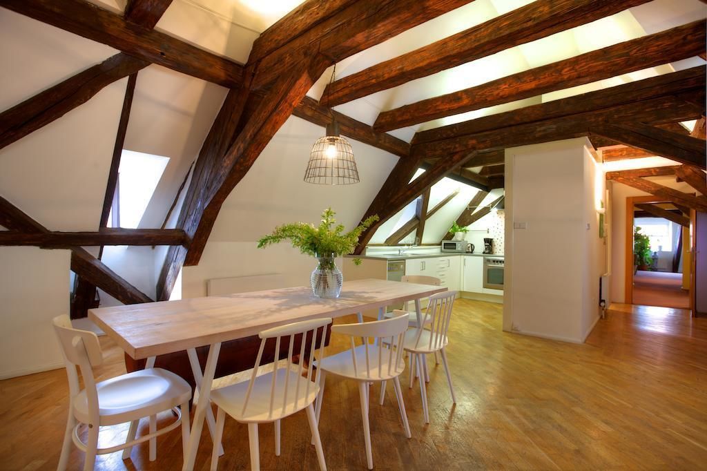 Uma sala de jantar com uma mesa de madeira e cadeiras brancas, um vaso com plantas verdes e vigas de madeira aparente.