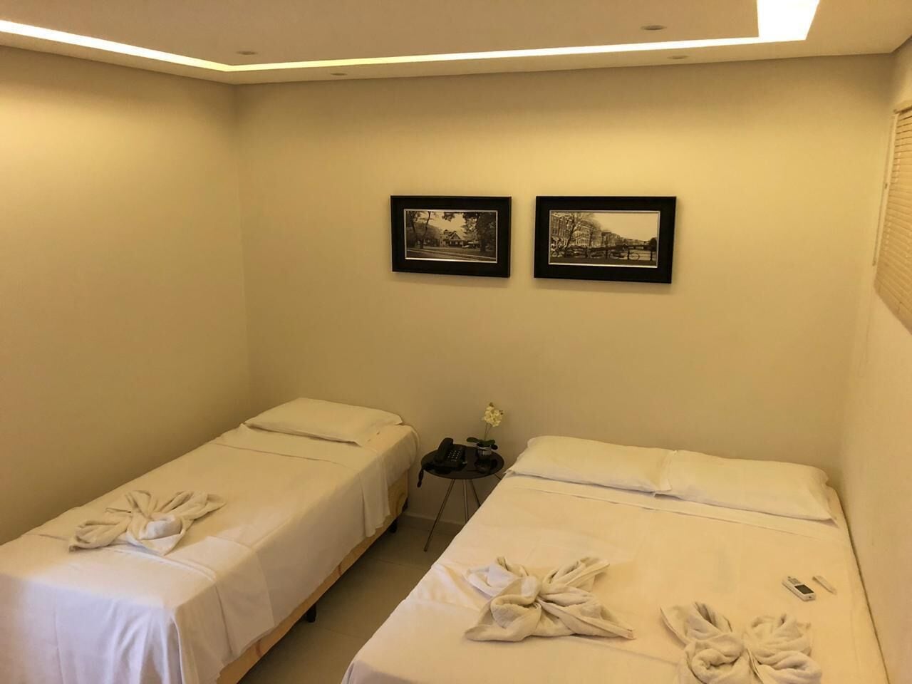 Quarto pequeno e limpo com duas camas de solteiro, uma mesinha de cabeceira com telefone e uma pequena planta, e duas quadros na parede.