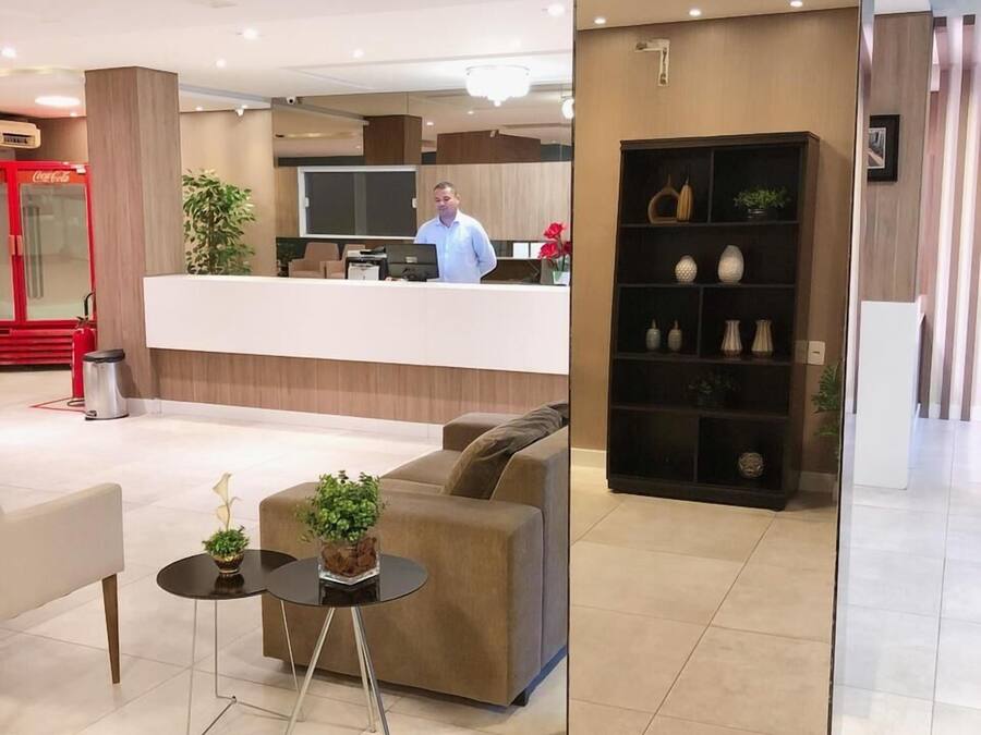 Um lobby moderno de hotel com uma receção, área de estar com um sofá e duas mesinhas, e uma estante decorativa.