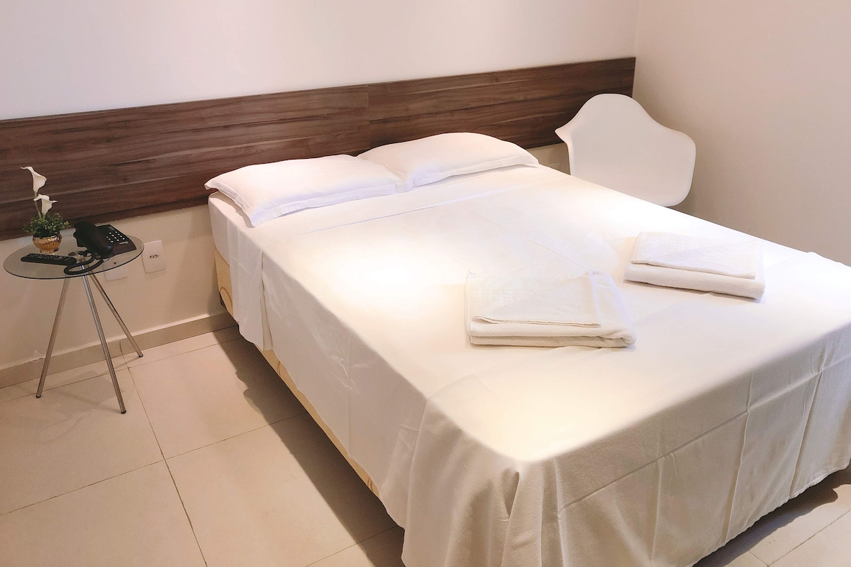 Uma cama bem arrumada com lençóis brancos, uma mesinha com um vaso e controles remotos, e uma cadeira encostada na parede.
