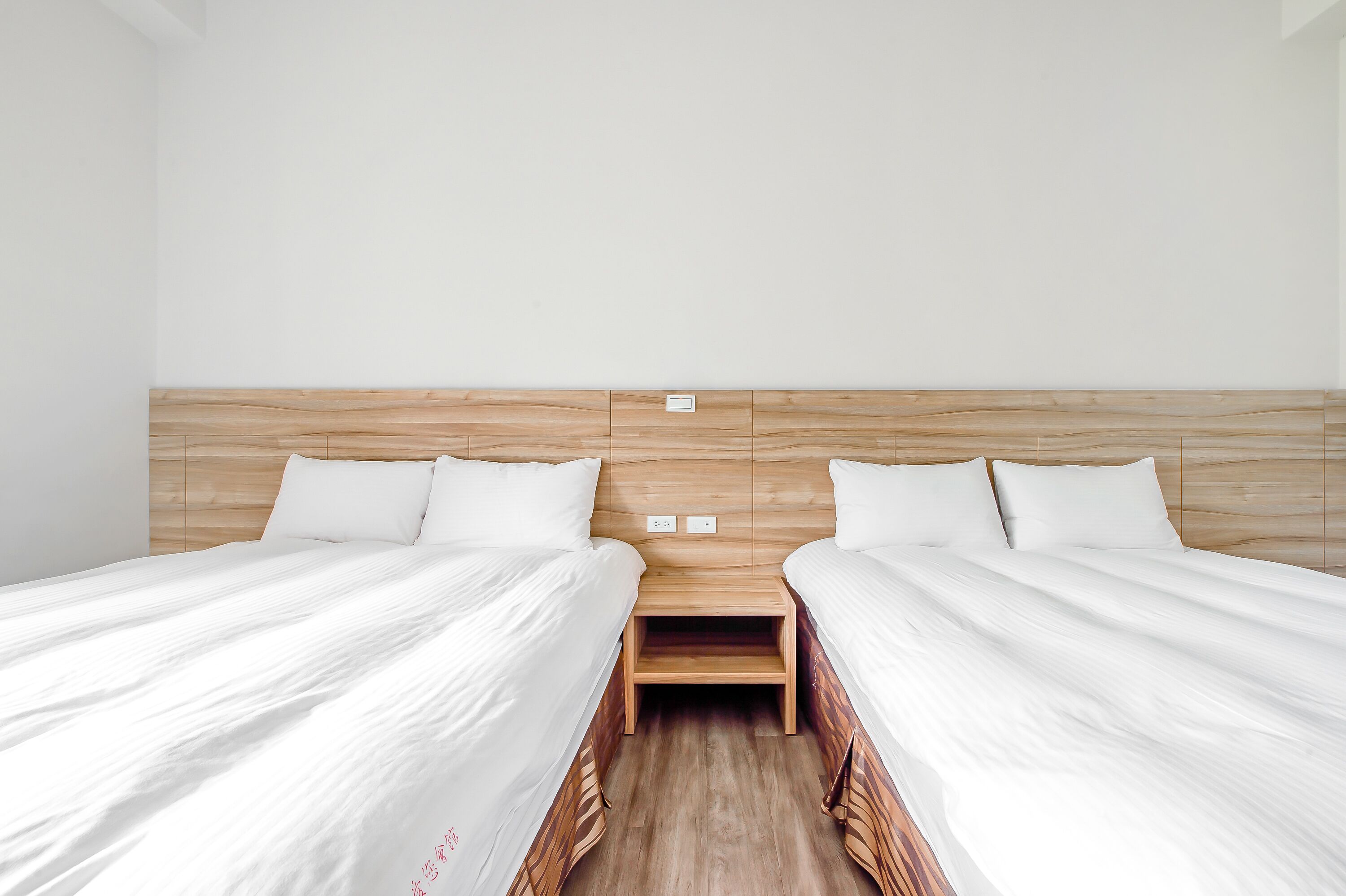 Quarto duplo com duas camas, uma cabeceira de madeira e um pequeno criado-mudo de madeira entre elas.