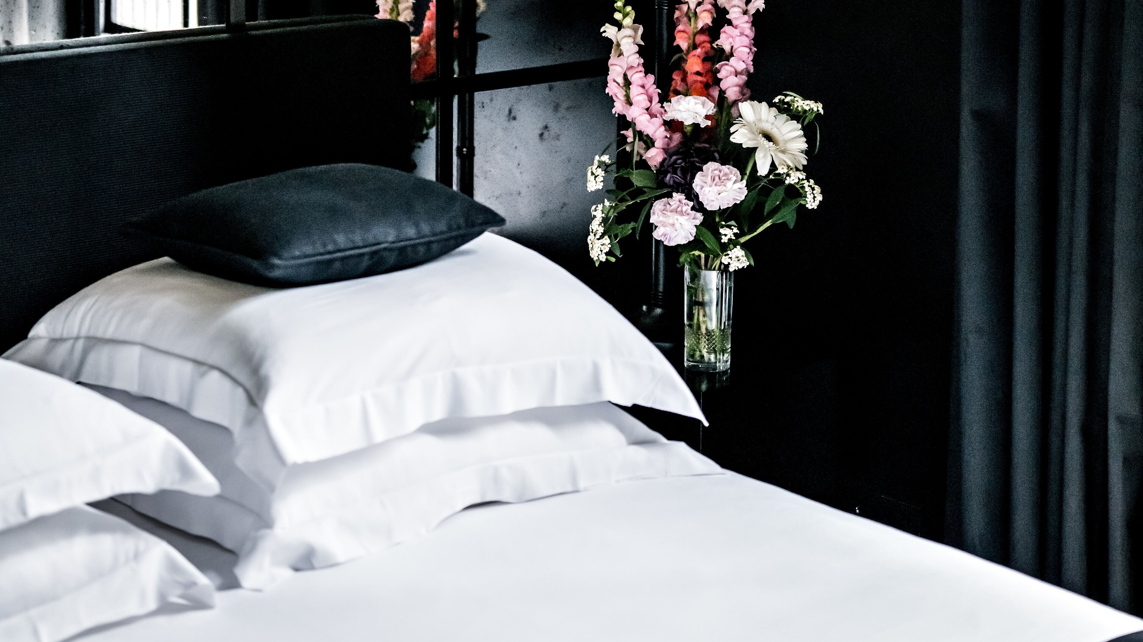Cama bem arrumada com lençóis brancos e almofada escura, um vaso com flores em uma mesa de cabeceira e uma cortina escura ao fundo.