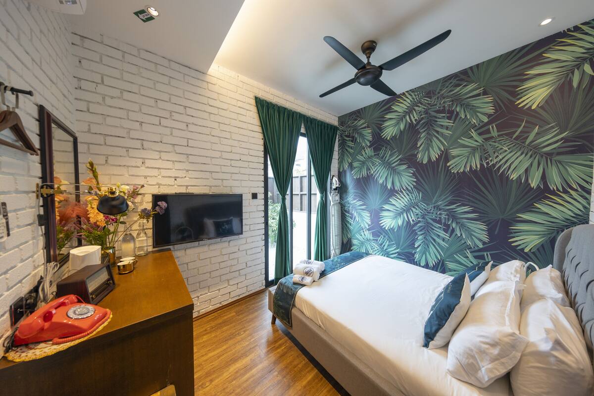 Quarto de hotel com parede de tijolos, cama com roupa de cama branca, ventoinha no teto, televisão e janela com cortinas verdes.