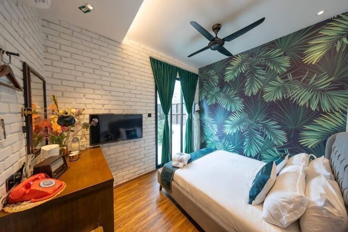 Quarto com cama, escrivaninha, televisão, ventoinha de teto e parede de tijolos.