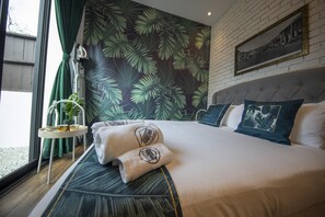 Quarto de hotel com uma cama grande, vista para o exterior, planta verde em uma mesa lateral e parede com padrão de folhagem tropical.