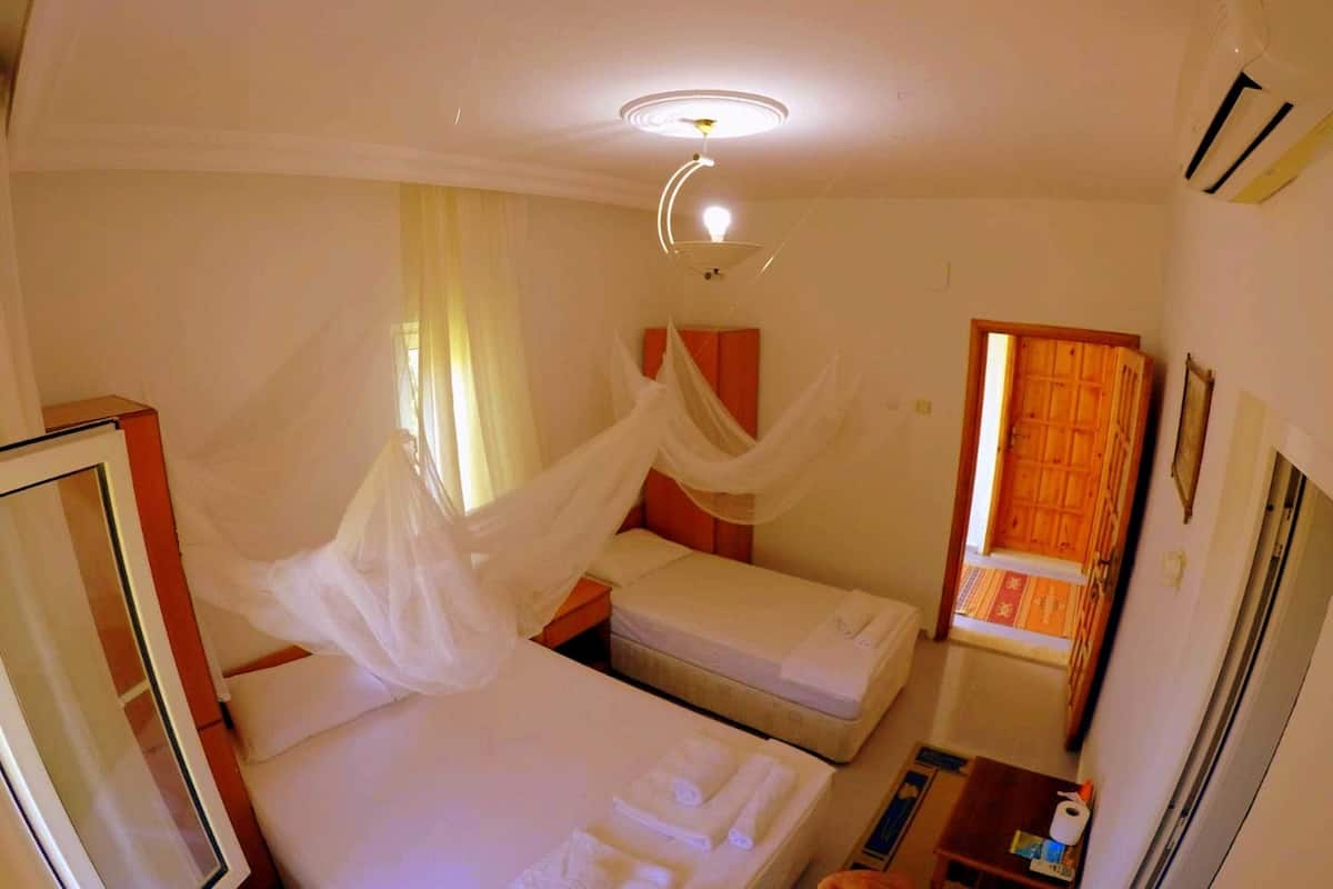 Quarto de hotel com duas camas, uma luminária no teto, uma porta de madeira e uma mesinha com uma garrafa e um lenço.