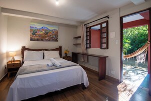 Quarto com cama, mesa de cabeceira, pintura, janela com vista para vegetação e rede do lado de fora.