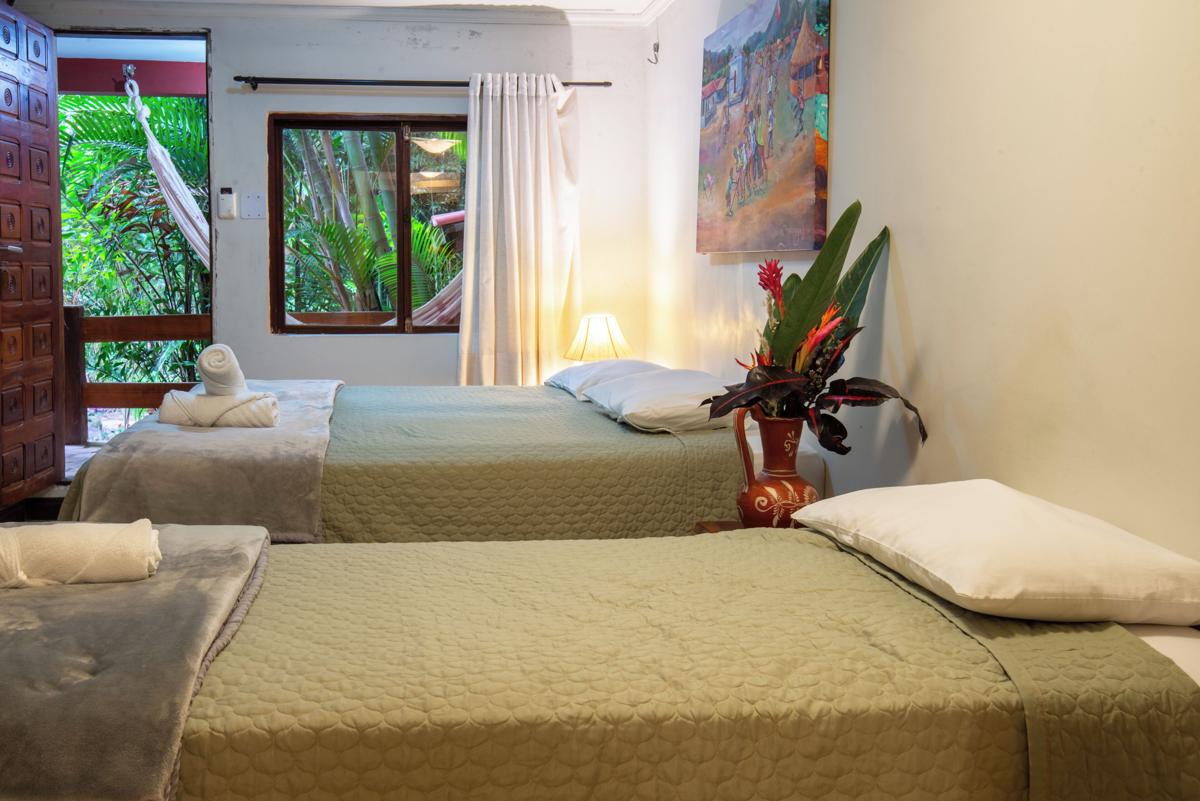 Quarto de hotel com duas camas, uma janela com cortinas, um quadro na parede e um vaso de plantas.