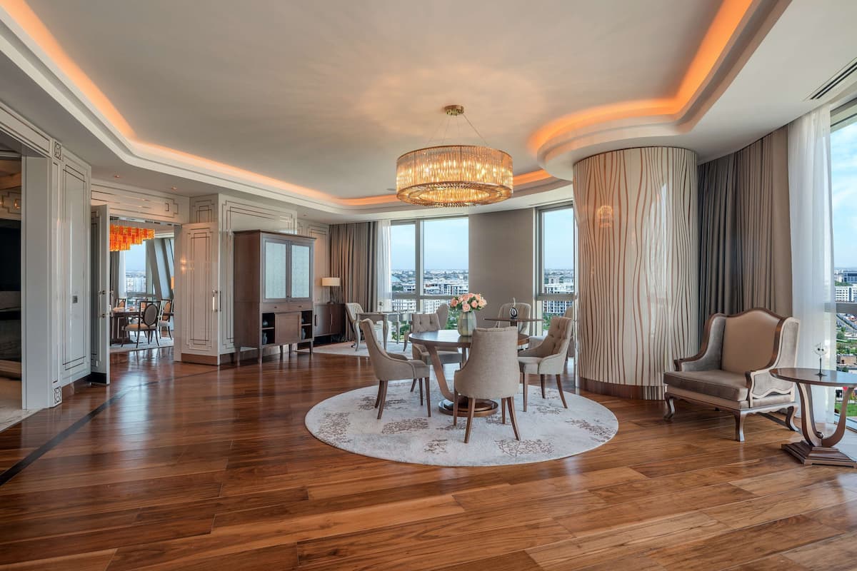 Uma sala de estar ampla com uma mesa de jantar redonda, assentos confortáveis, um lustre e grandes janelas com vista para a cidade.