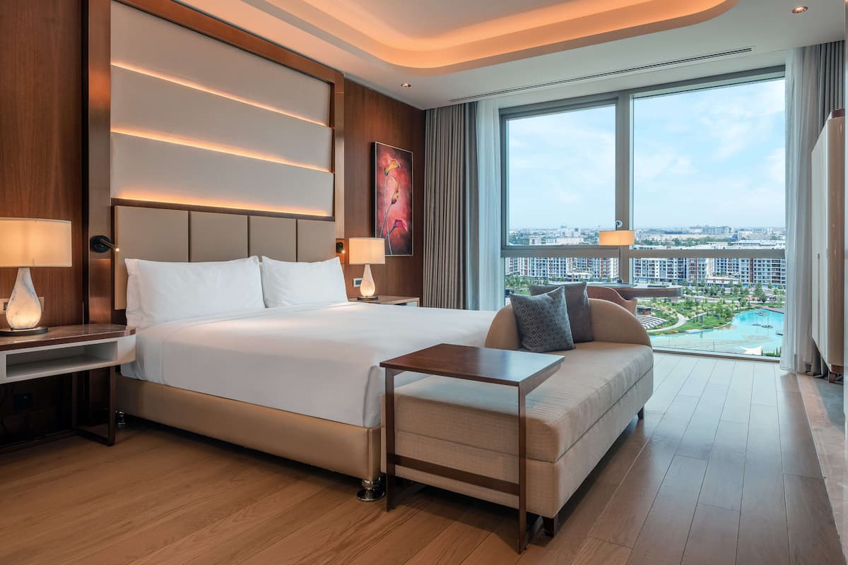 Quarto de hotel moderno com uma cama grande, um sofá, uma mesinha e vista para a cidade através de janelas amplas.
