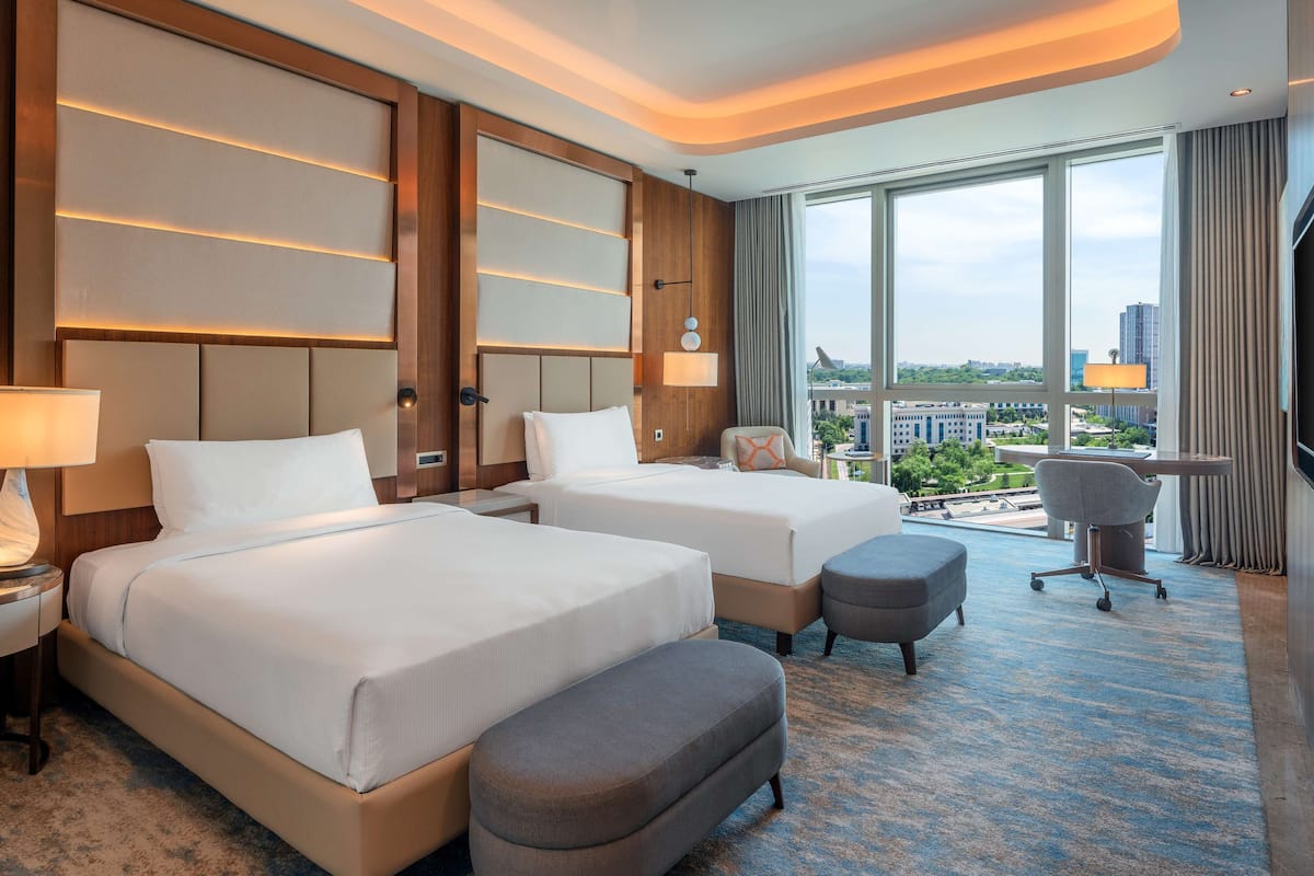 Quarto de hotel com duas camas, uma escrivaninha, uma cadeira e uma janela grande com vista para a cidade.