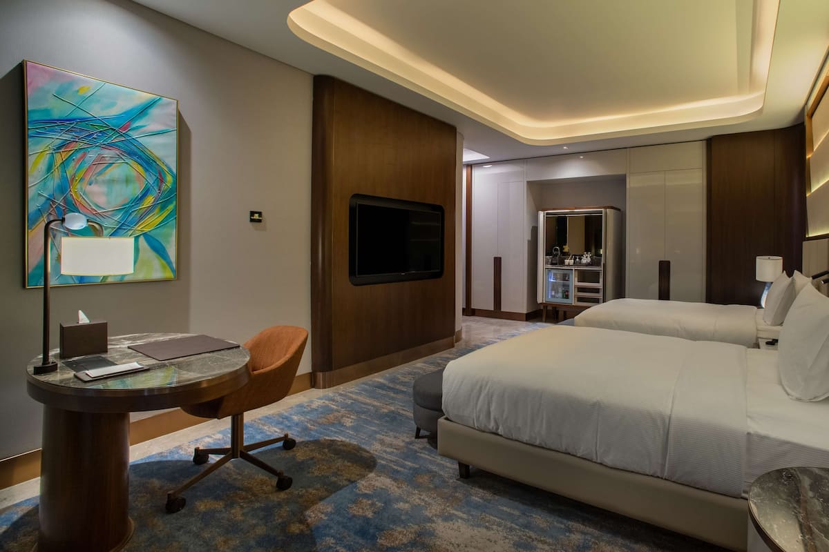 Quarto de hotel com uma cama, uma escrivaninha com cadeira, uma televisão na parede e um quadro abstrato grande.