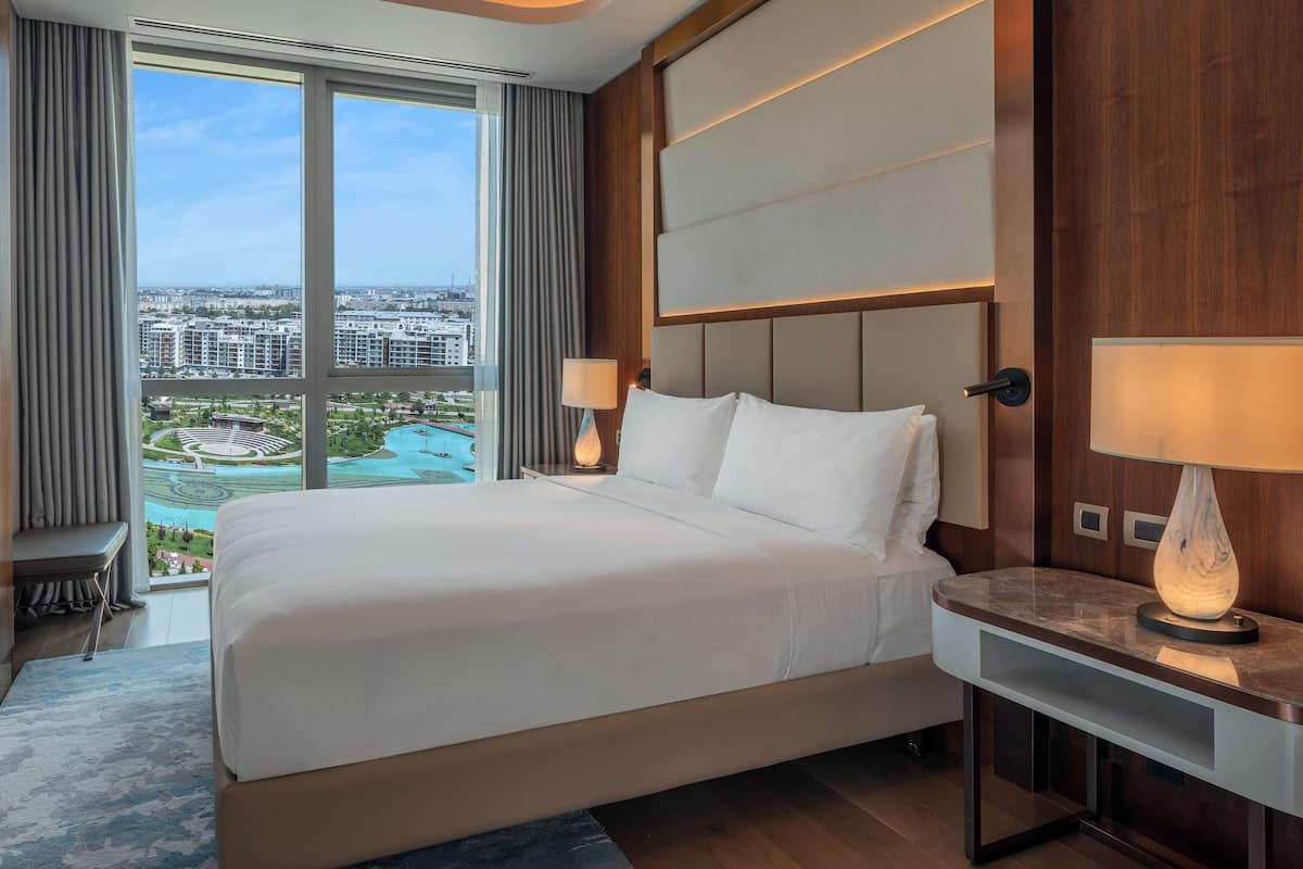 Quarto de hotel com uma cama grande, duas mesas de cabeceira com abajures, vista para a cidade e uma piscina através de uma janela ampla.