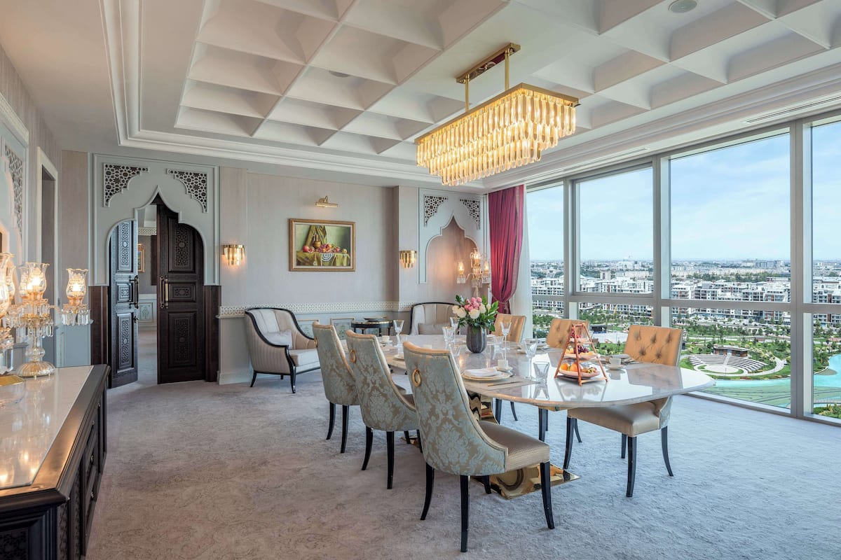 Uma ampla área de jantar com uma mesa grande, cadeiras elegantes e um lustre. Uma vista da cidade é visível através de grandes janelas.