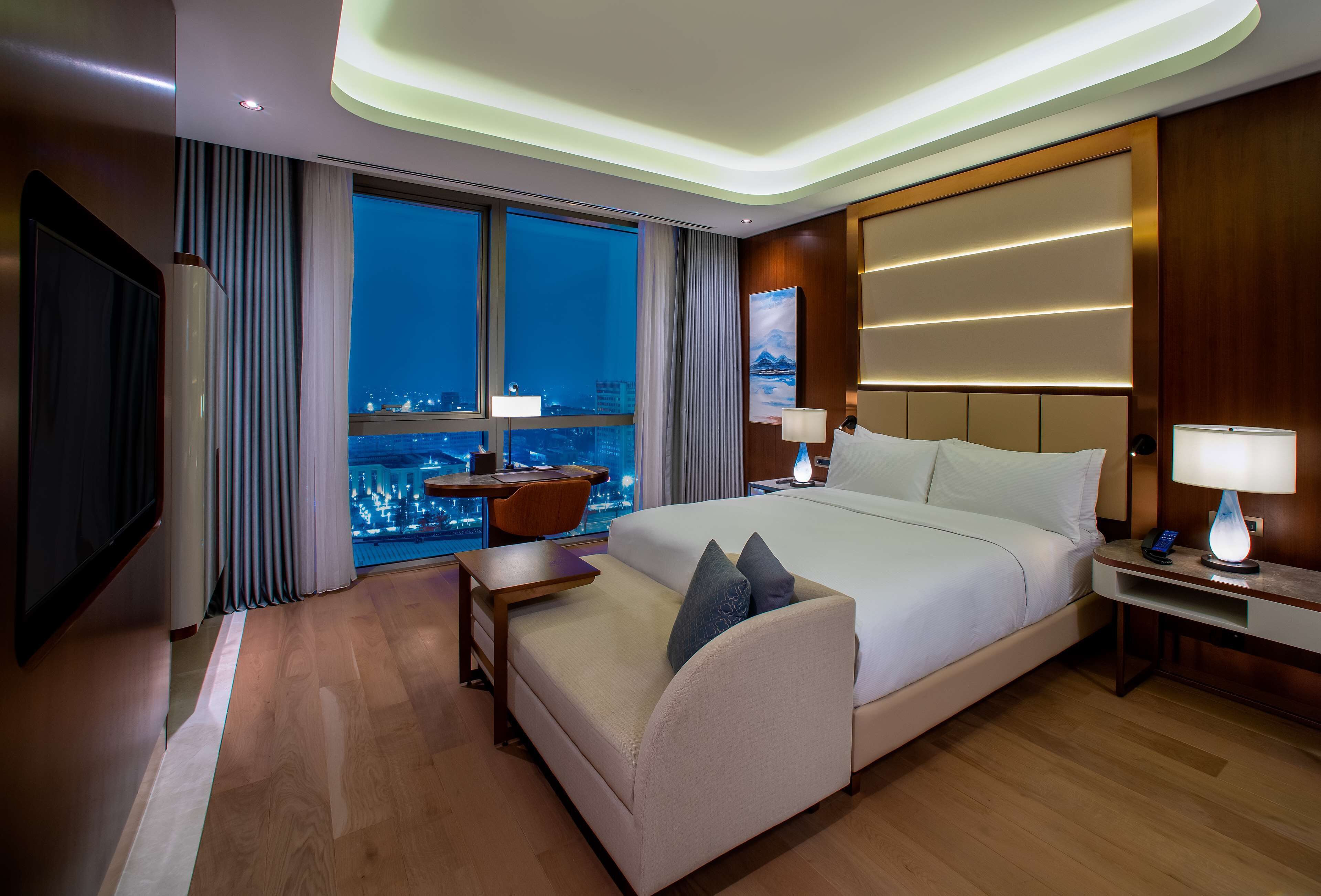 Quarto de hotel moderno com uma cama grande, uma escrivaninha e vista para a cidade à noite.