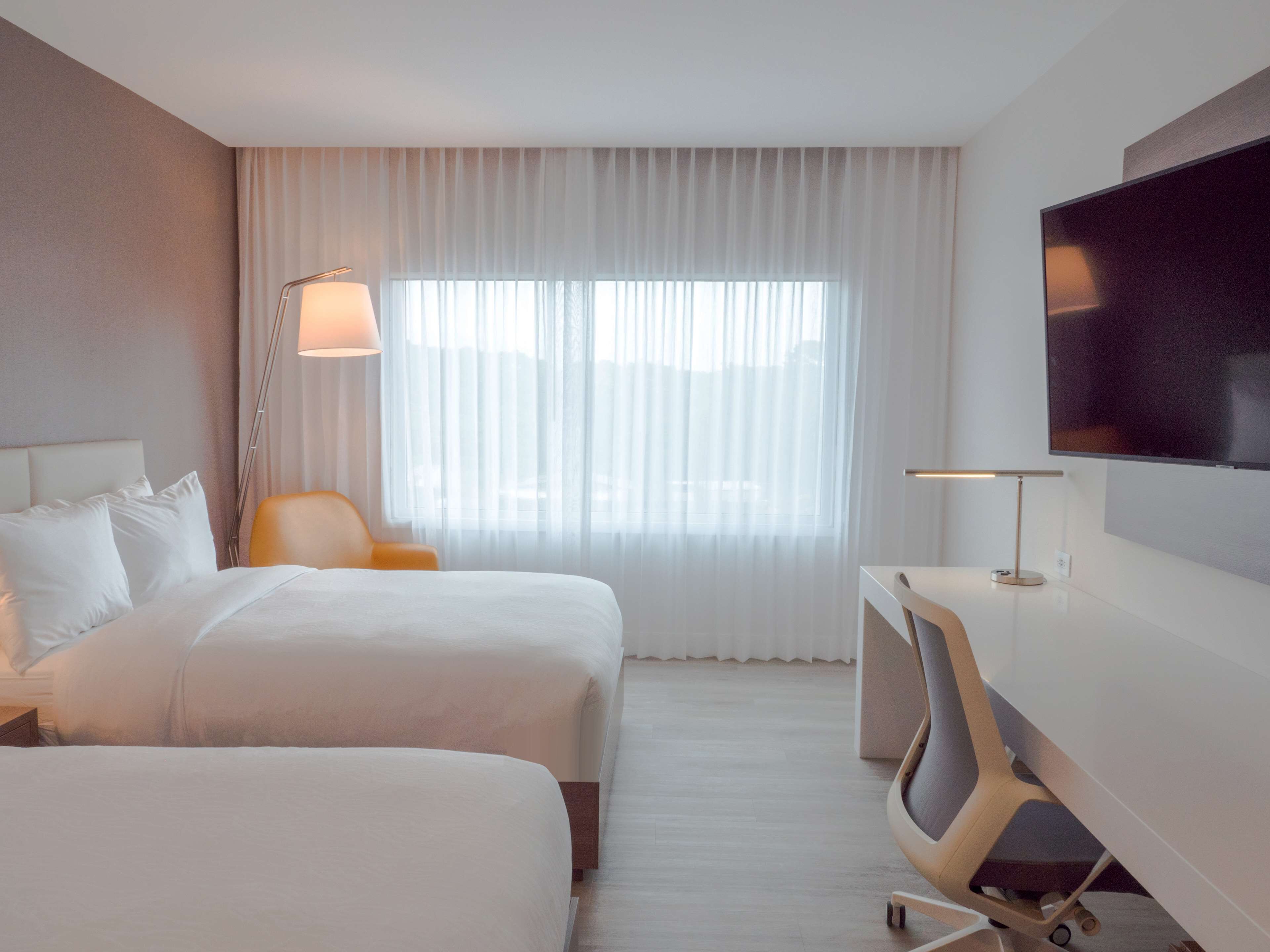 Quarto de hotel com duas camas, uma escrivaninha, uma cadeira, uma TV e uma janela grande com cortinas leves.