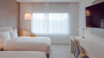Quarto de hotel com duas camas, uma escrivaninha, uma cadeira, uma TV e uma janela grande com cortinas leves.
