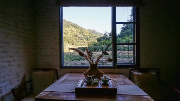 Quarto com uma grande janela, uma mesa de madeira com um vaso e chávenas, e uma parede de tijolo.