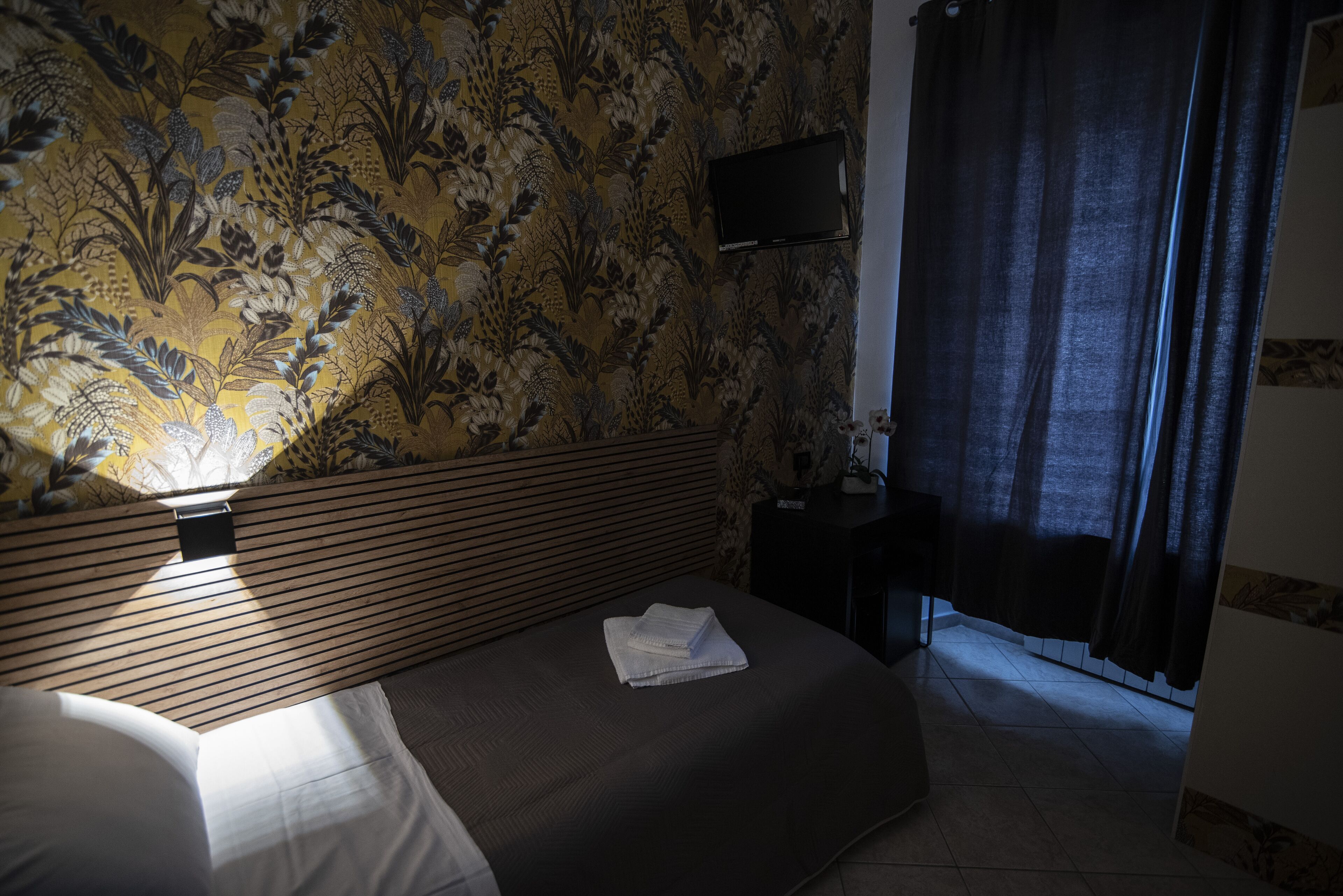 Quarto de hotel com cama, mesa de cabeceira, televisão e cortina.