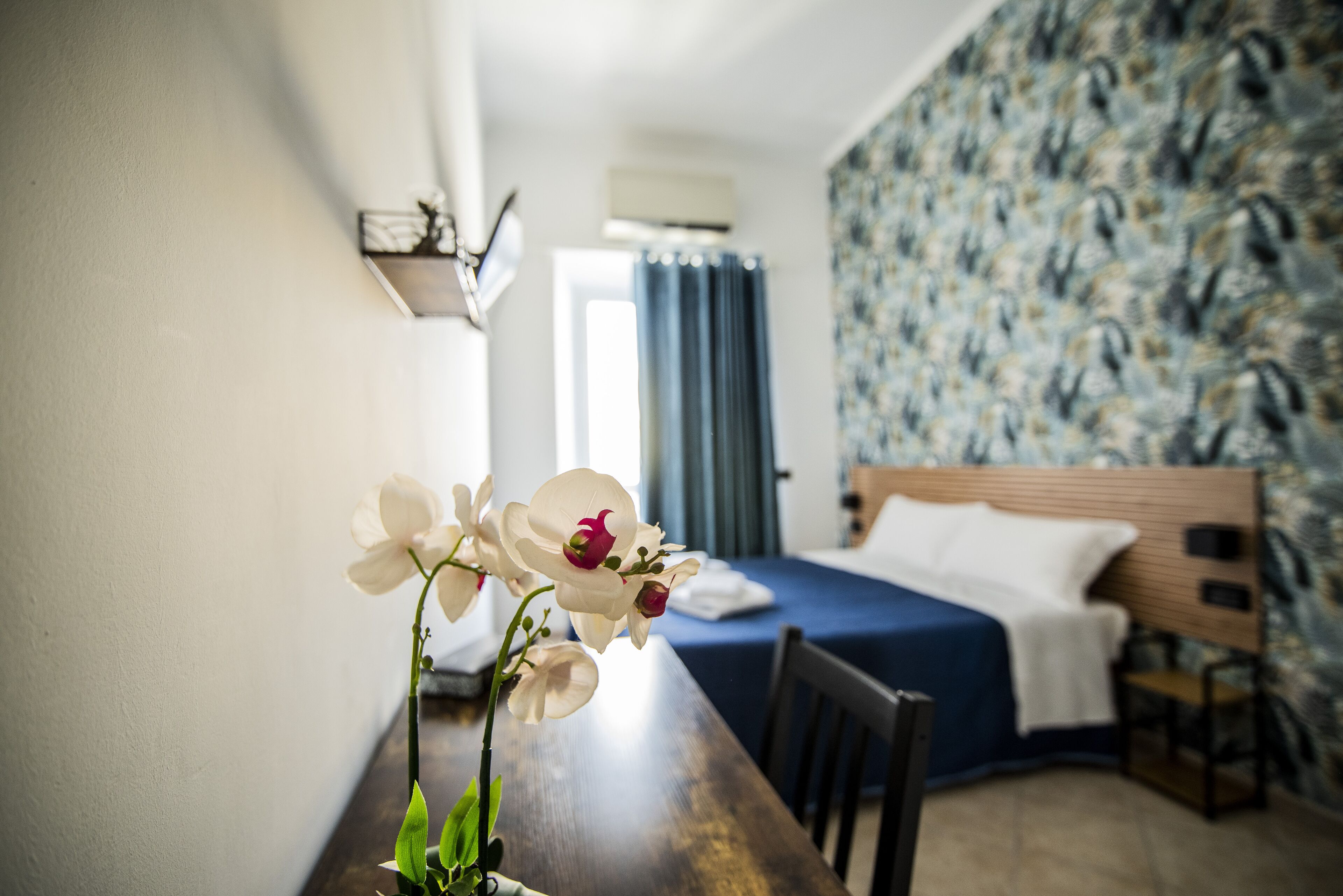 Quarto de hotel com cama de madeira, colcha azul, mesa de cabeceira de madeira e papel de parede floral.
