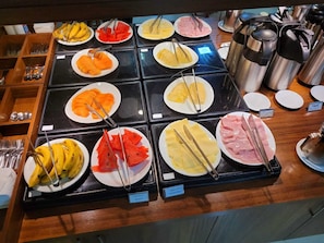 Buffet com diversas opções de comida, como bananas, melancia, queijo e presunto.