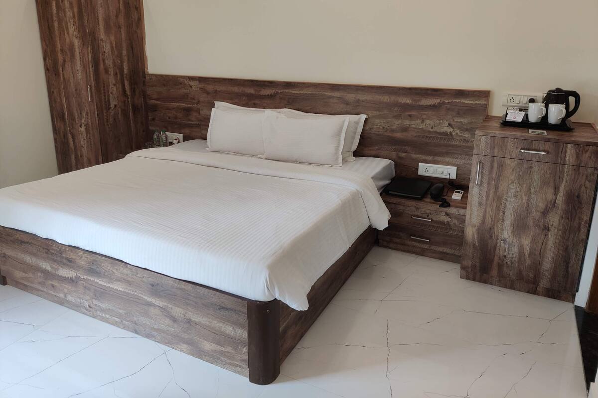 Cama bem arrumada com cabeceira de madeira, mesa de cabeceira com telefone e chaleira, e pequeno armário com gavetas.