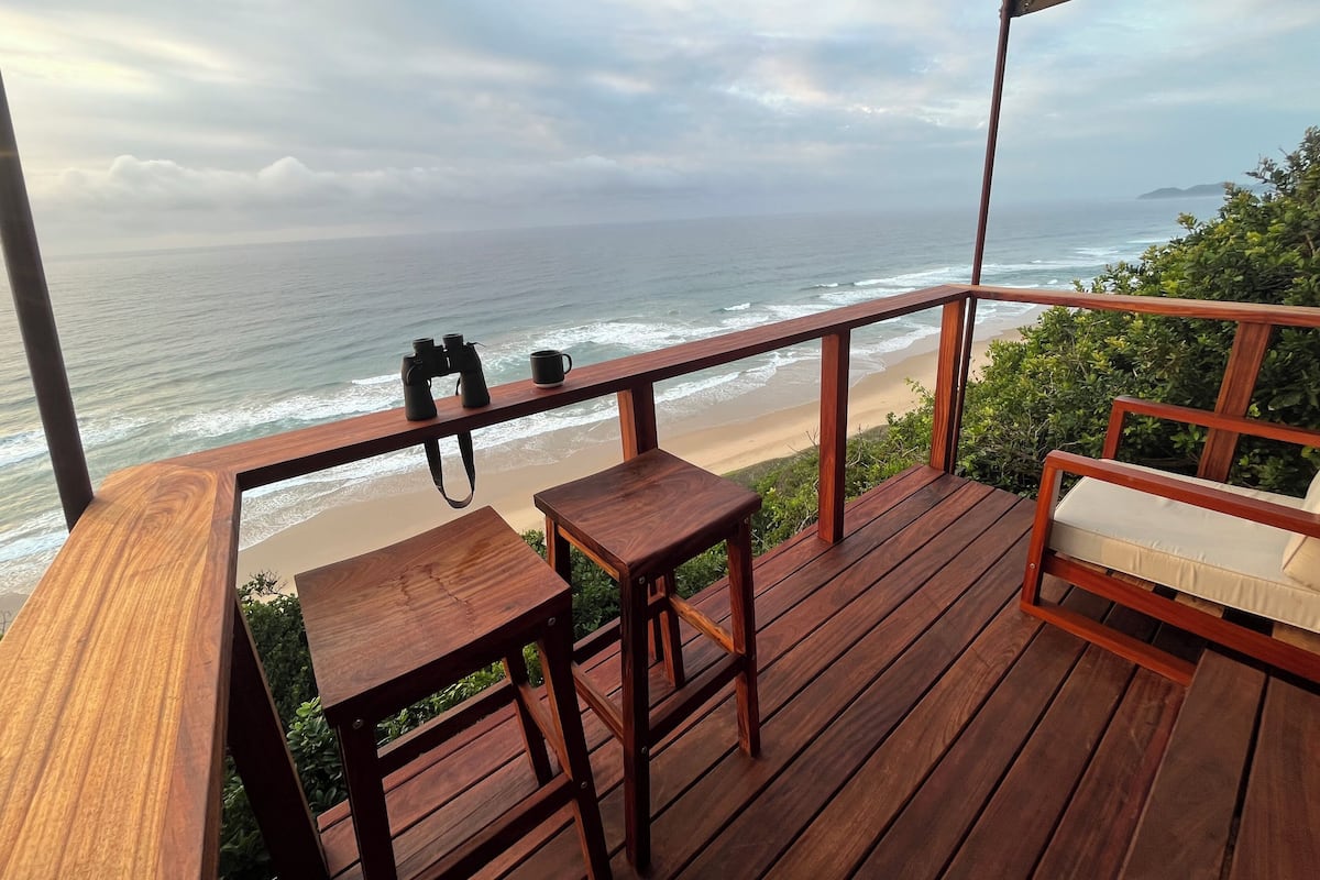 Um deck de madeira com vista para o oceano, um par de binóculos e um banco.