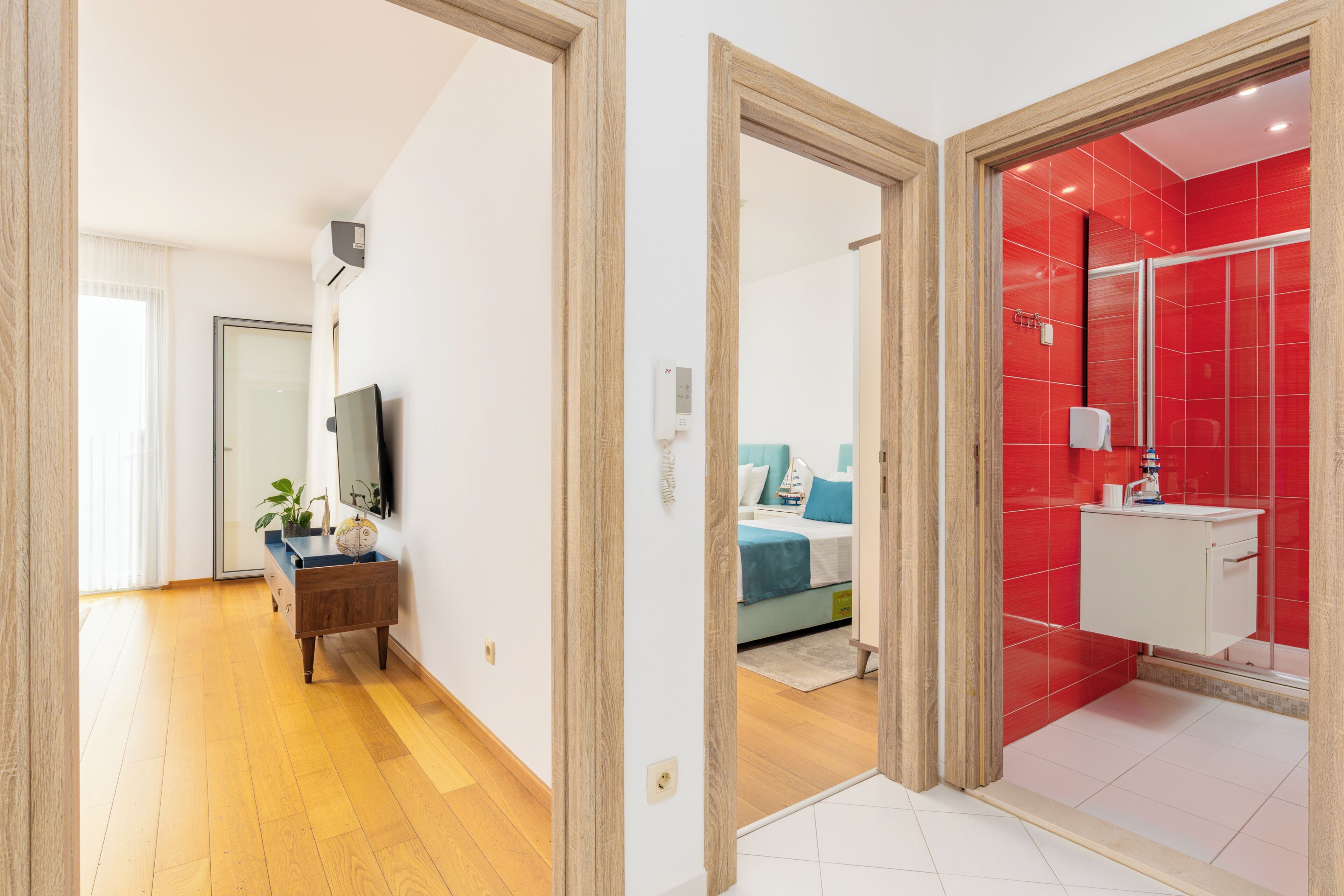Apartamento moderno com piso de madeira, televisão, planta, cama e banheiro vermelho.
