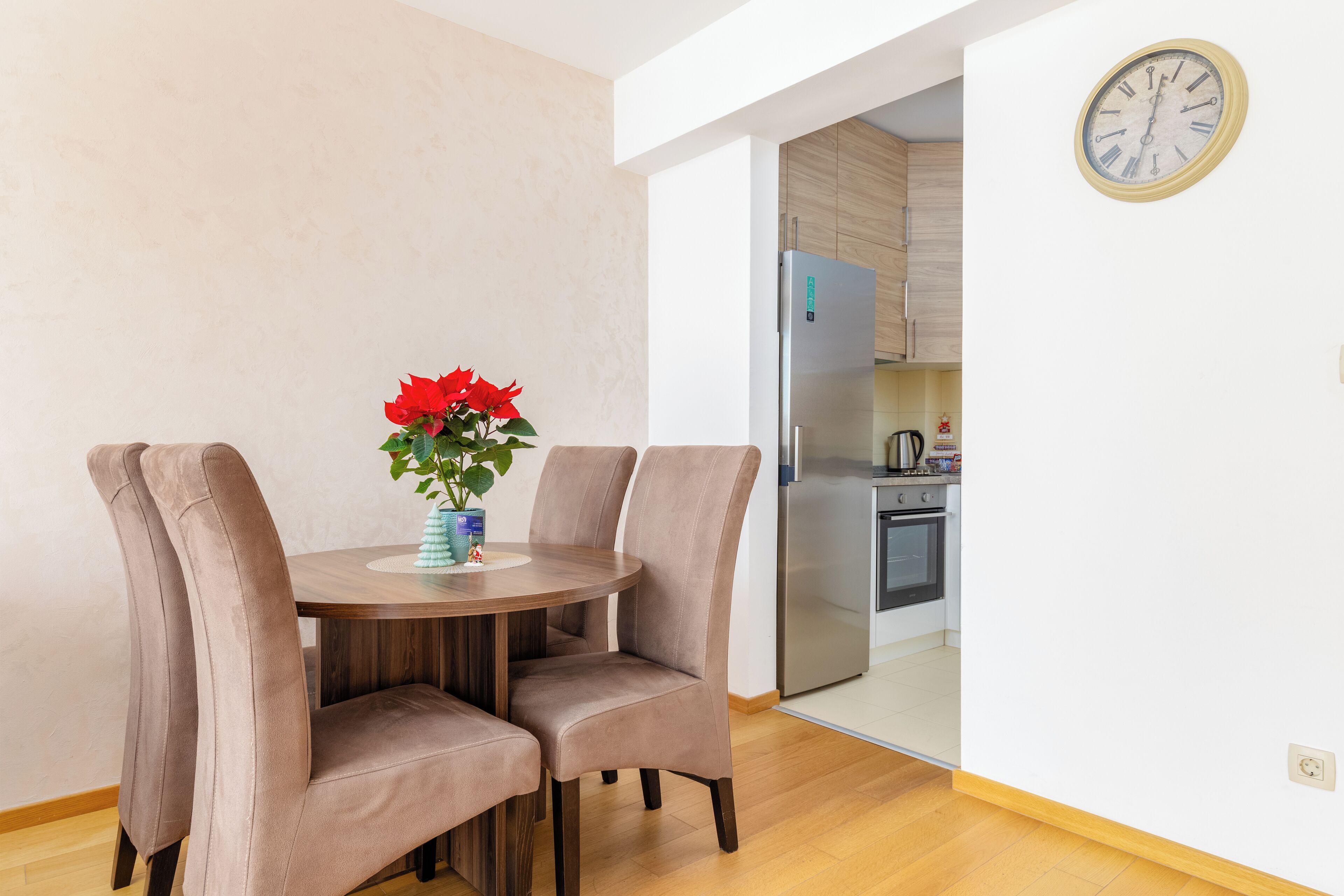 Uma sala de jantar com uma mesa redonda, duas cadeiras estofadas e um vaso com flores vermelhas.