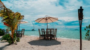 Uma praia com mesa e cadeiras sob guarda-sóis, uma palmeira e um lampião.