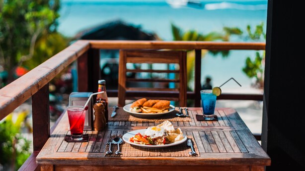 Uma mesa de madeira com uma refeição, bebidas e vista para o mar.