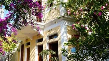 Um edifício de dois andares com telhado vermelho, paredes brancas e uma varanda. Escadas levam à entrada, e o edifício é cercado por vegetação e plantas floridas.