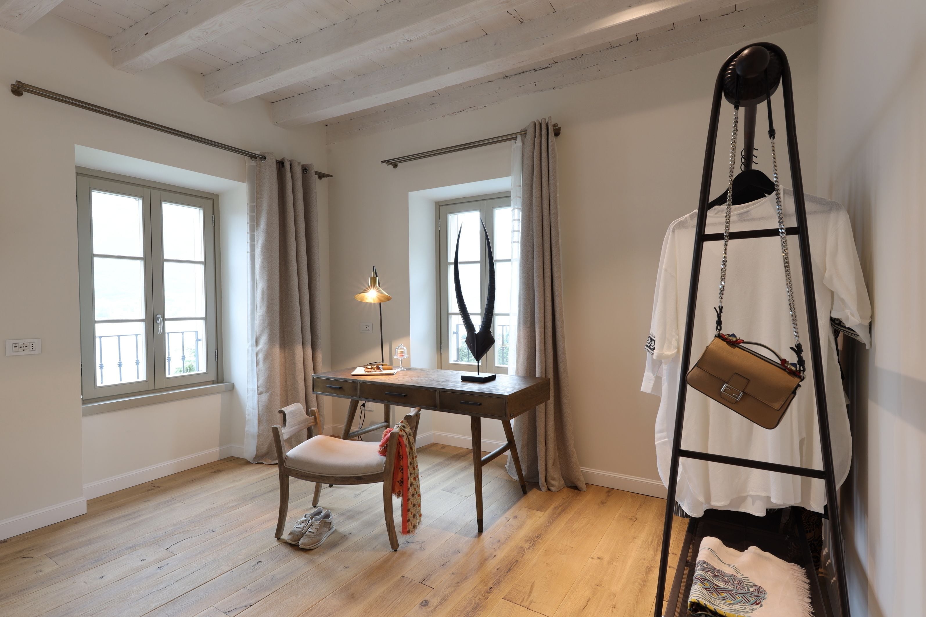 Quarto com chão de madeira, uma escrivaninha com abajur, uma cadeira, um cabideiro com roupas e uma mala, e duas janelas com cortinas.