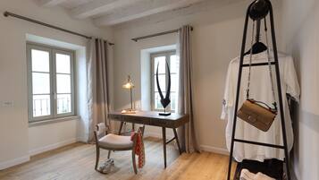 Quarto com chão de madeira, uma escrivaninha com abajur, uma cadeira, um cabideiro com roupas e uma mala, e duas janelas com cortinas.