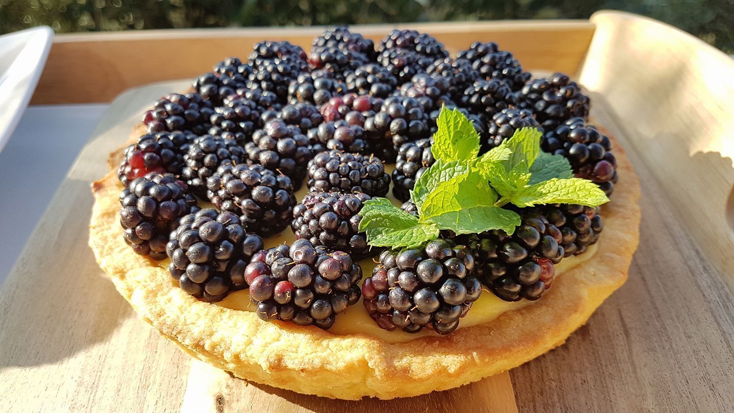 Uma torta redonda coberta com amoras e folhas de hortelã, sobre um tabuleiro de madeira.
