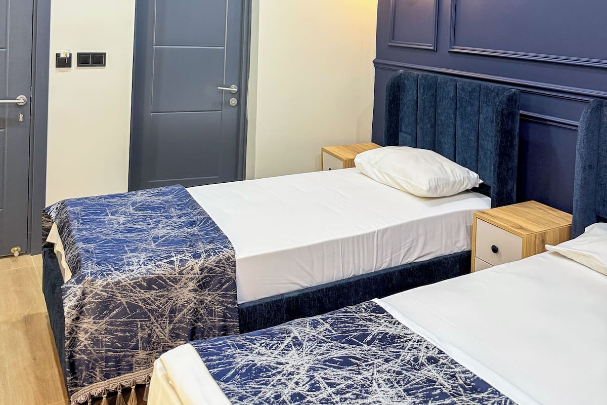Quarto de hotel com duas camas, cabeceira azul escura, mesa de cabeceira de madeira pequena e parede de cor clara.