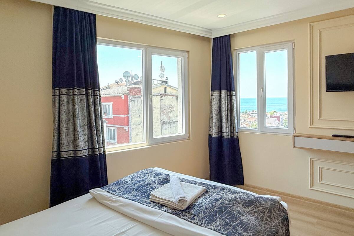 Quarto de hotel com cama, duas janelas com cortinas, televisão fixada na parede e vista para edifícios e o mar.
