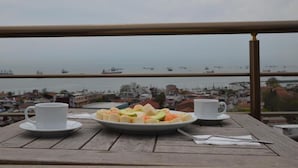 Mesa de varanda com um prato de frutas e duas xícaras brancas, com vista para uma paisagem urbana e um corpo de água com navios.