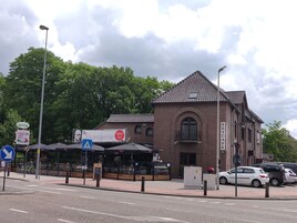 Um edifício com telhado de azulejos e janelas em arco, ao lado de uma rua com carros estacionados e um café com mesas ao ar livre.
