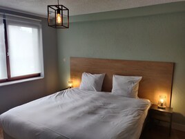 Cama bem arrumada em quarto com cabeceira de madeira, duas luminárias de apoio e janela com persianas.