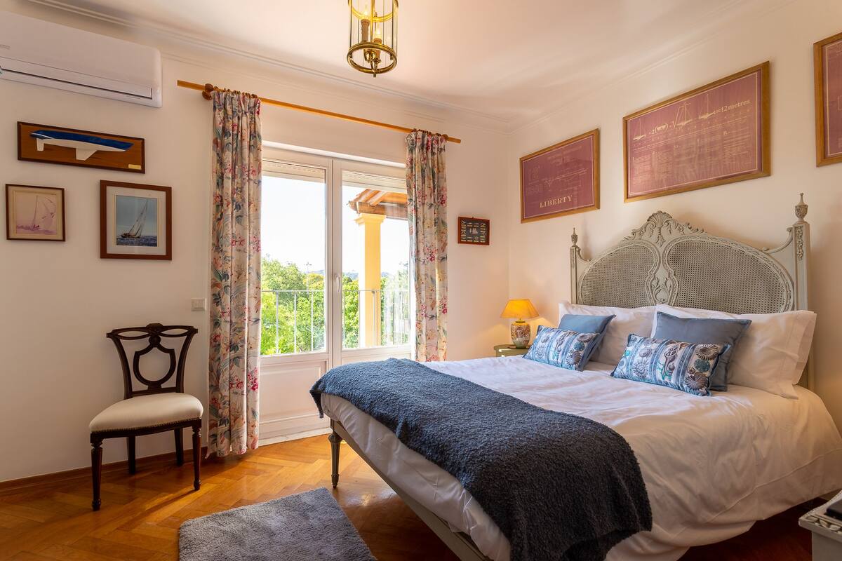 Quarto com cama grande, uma cadeira, janela com cortinas e quadros na parede.